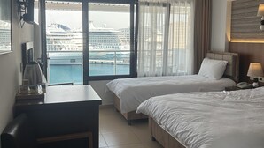 Quarto de hotel com duas camas, uma mesa, uma cadeira, uma luminária e vista para um navio de cruzeiro e para a água.