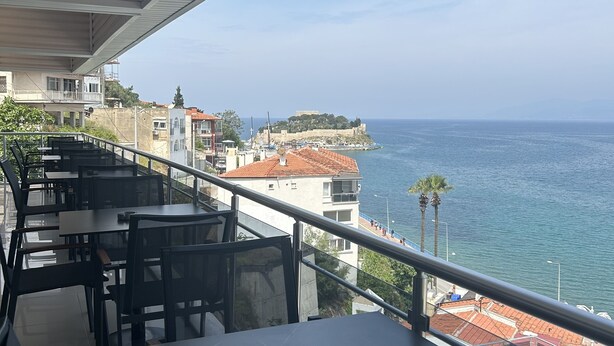 Uma área de estar externa com mesas e cadeiras, com vista para a costa, edifícios e uma fortaleza numa colina.