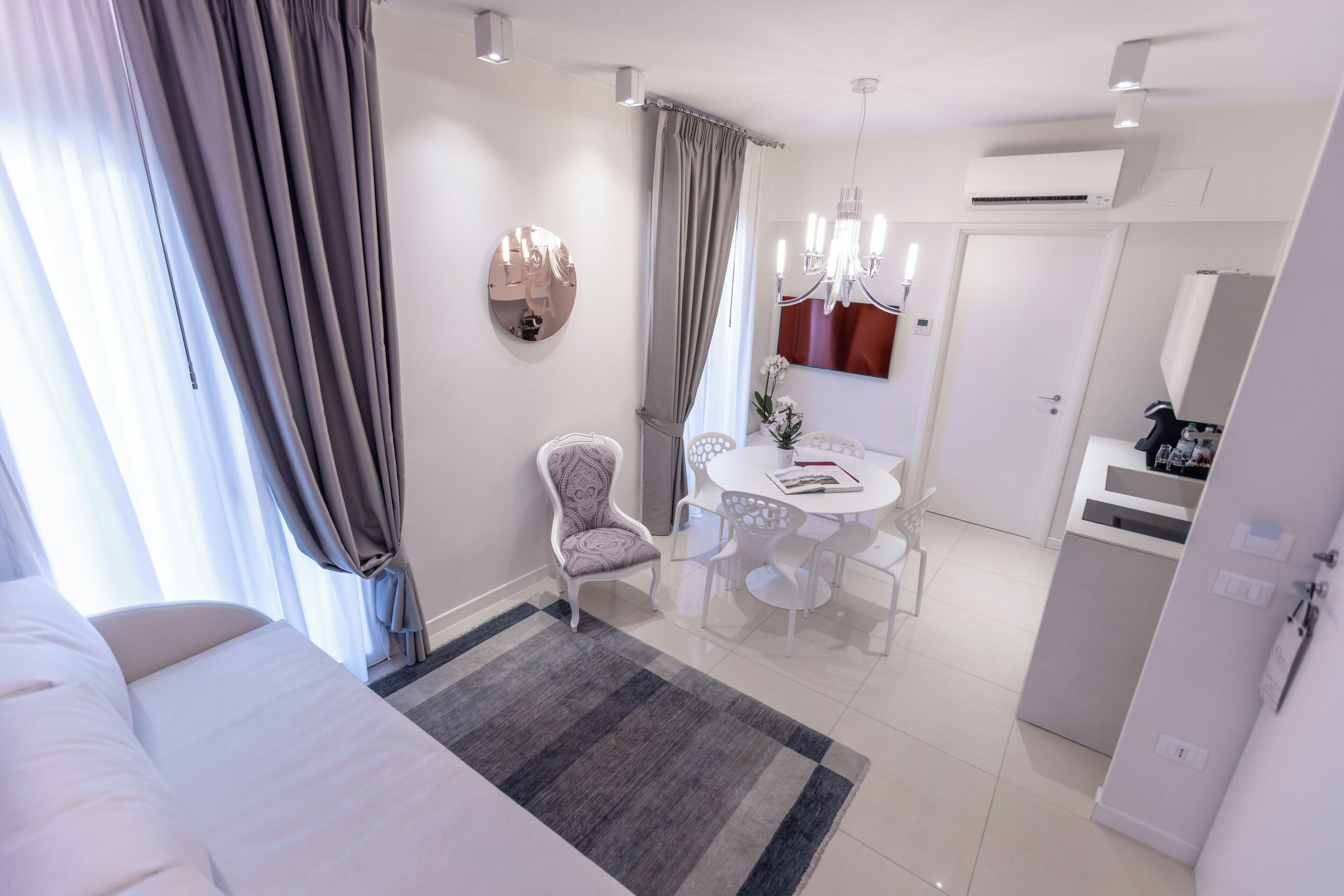 Quarto de hotel moderno com uma cama, área de jantar com mesa e cadeiras, um candelabro e um aparelho de ar condicionado.