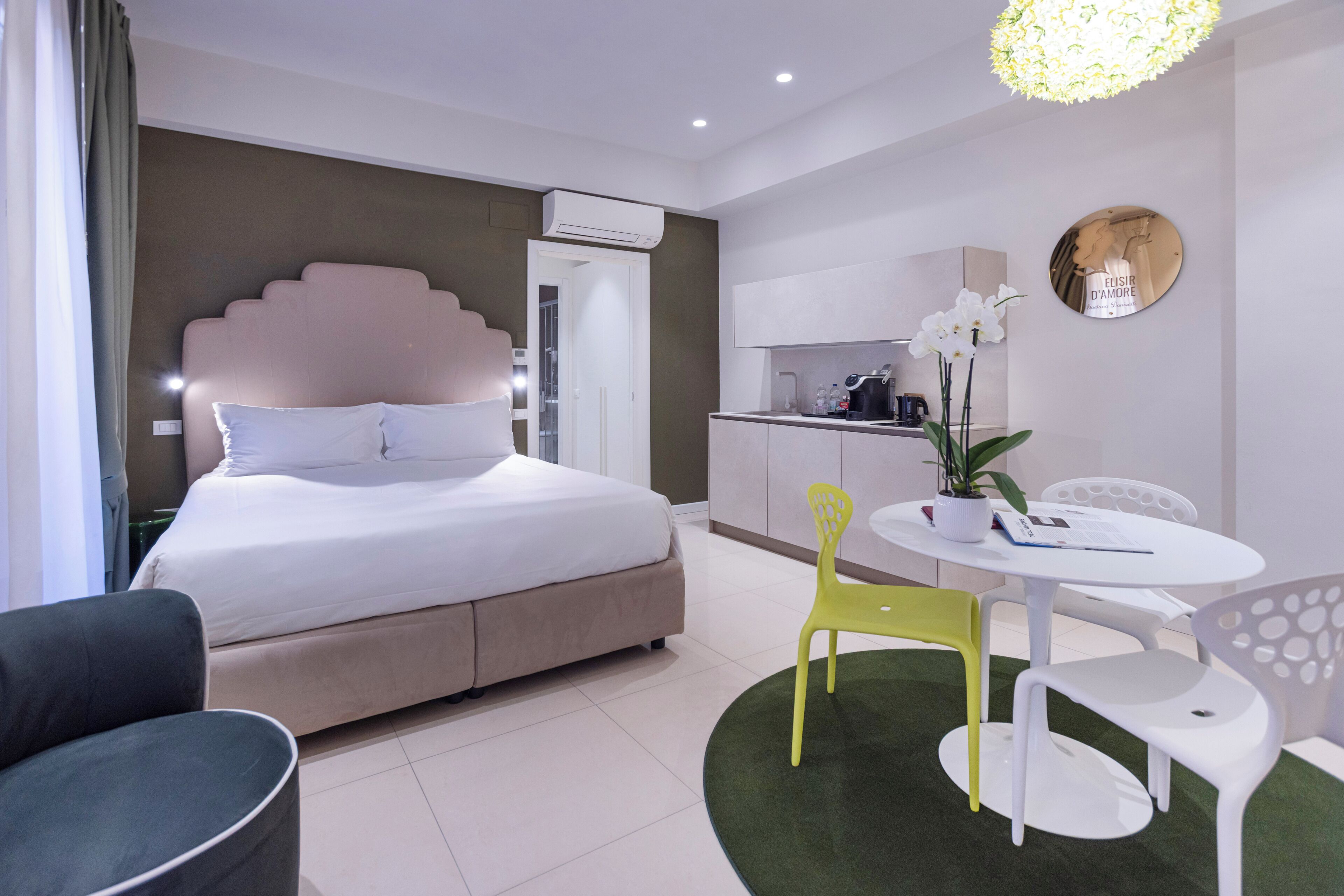 Quarto de hotel com cama, pequeno kitchenette, mesa de jantar redonda com cadeiras e espelho na parede.