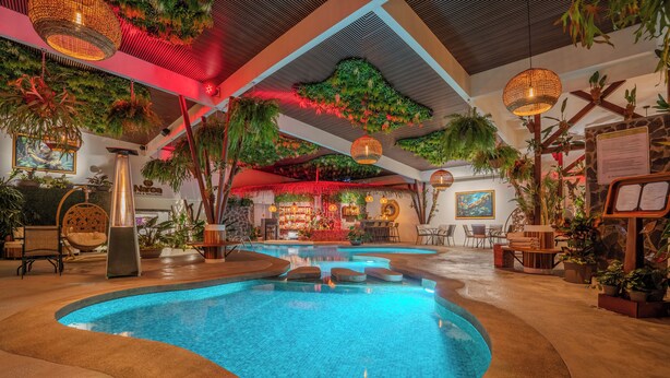 Uma área interna com piscina com tema tropical, incluindo uma piscina grande, áreas de estar e vegetação exuberante.