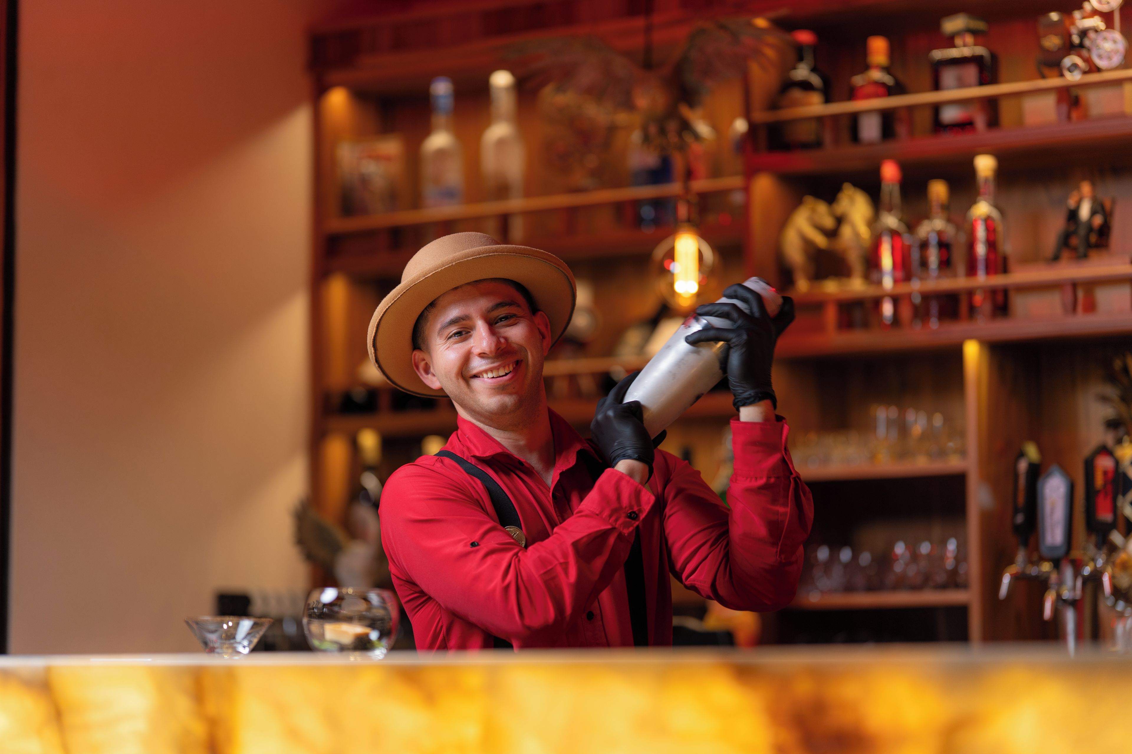 Um bartender com camisa e chapéu vermelhos, preparando uma bebida atrás do balcão, com prateleiras cheias de garrafas.