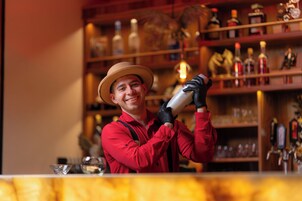 Um bartender com camisa e chapéu vermelhos, preparando uma bebida atrás do balcão, com prateleiras cheias de garrafas.