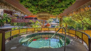 Uma banheira de hidromassagem rodeada por uma cerca decorativa, com um jardim exuberante e uma estrutura de telhado de palha ao fundo.