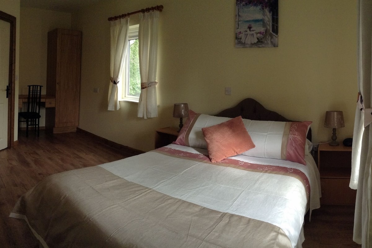 Um quarto com uma cama, uma mesa de cabeceira, uma luminária, uma janela com cortinas, uma porta e um quadro na parede.