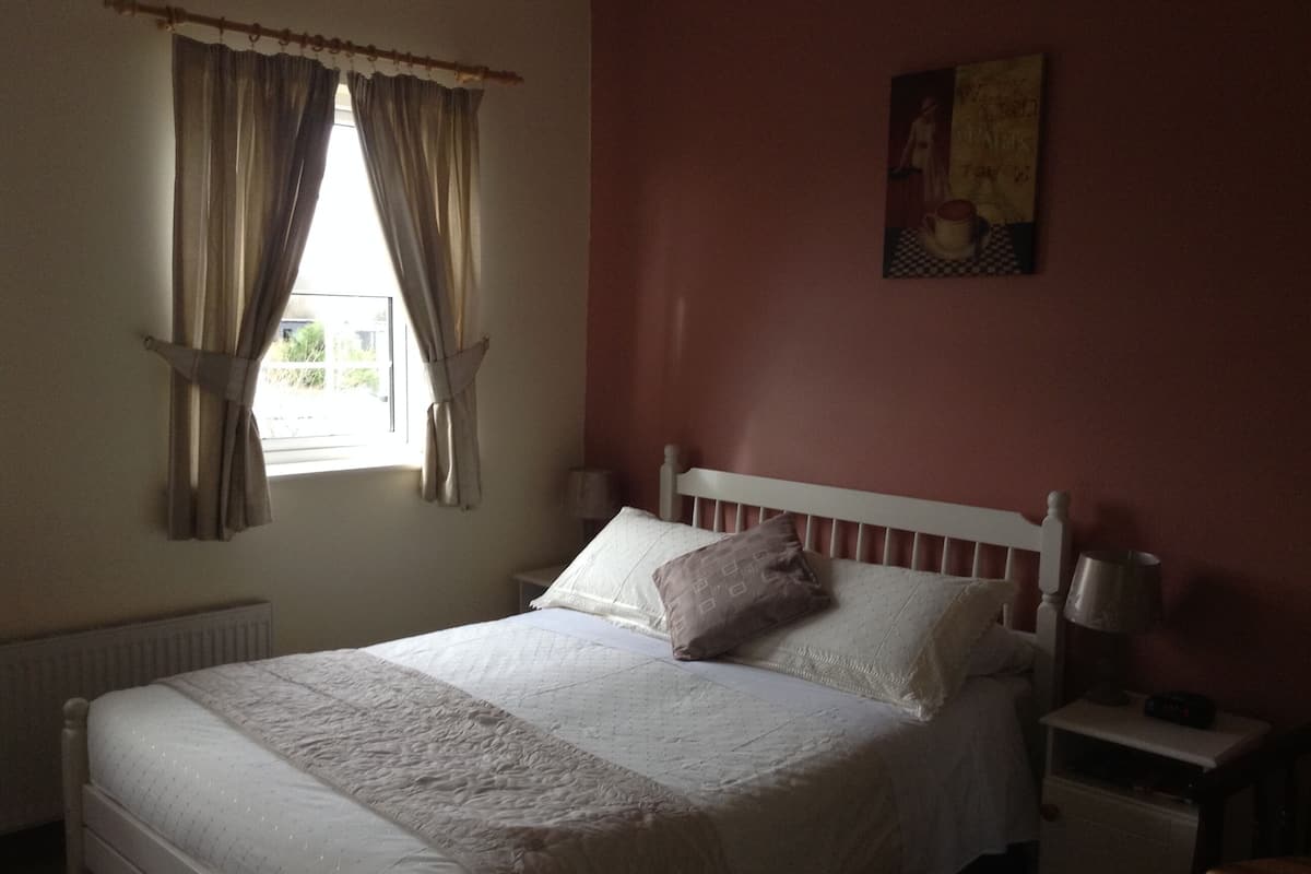 Quarto com cama, mesa de cabeceira, janela com cortinas e um quadro pendurado na parede.