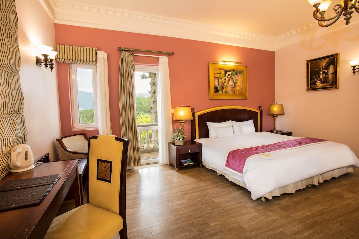 Quarto de hotel com cama, secretária com chaleira, cadeira, mesa de cabeceira com luminárias e janela com cortinas.