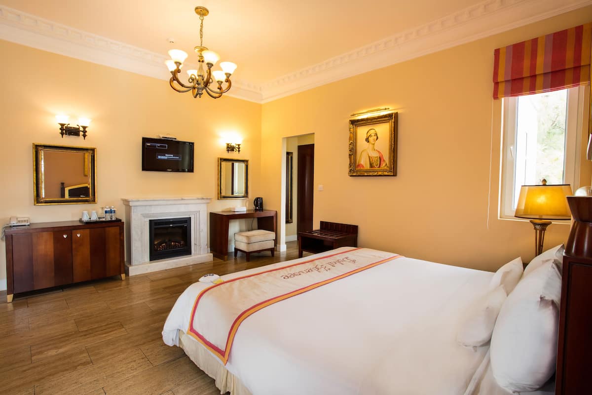 Quarto de hotel com uma cama grande, lareira, escrivaninha com cadeira, televisão e um quadro na parede.