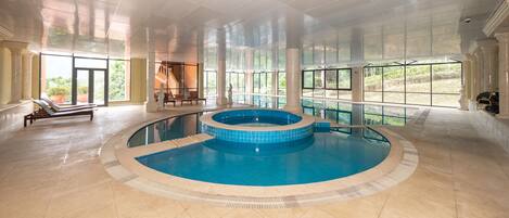 Uma grande piscina interior com uma banheira de hidromassagem, rodeada por amplas janelas e chão de pedra.