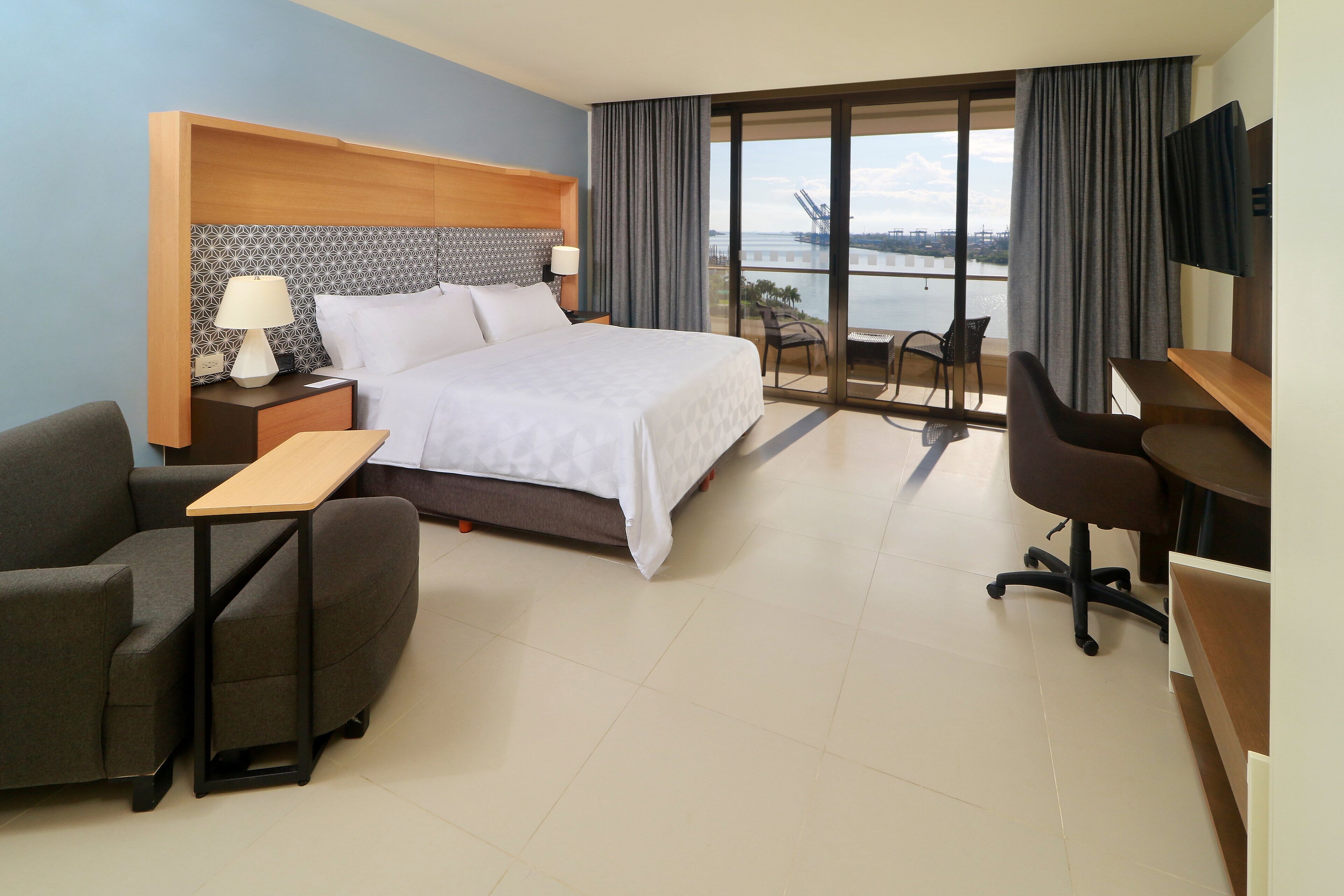 Quarto de hotel com cama, escrivaninha, cadeira e vista para um corpo de água.