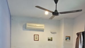 Uma cama de solteiro com base vermelha, roupa de cama branca e manta marrom. Há um ventilador de teto, dois aparelhos de ar condicionado instalados na parede e um quadro na parede.