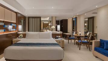 Quarto de hotel moderno com uma cama grande, um espaço de estar com sofá e cadeiras, uma mesa de jantar e um banheiro com espelho e toucador.