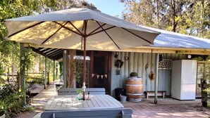 Um pátio externo com mesa e bancos sob um grande guarda-sol, cercado por vegetação e um edifício rústico com geladeira e barril.