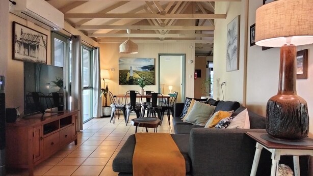 Sala de estar com sofá, TV e luminária. Há uma área de jantar com cadeiras e mesa.