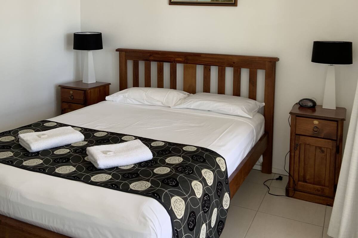 Uma cama bem arrumada com colcha estampada, duas mesas de cabeceira com abajures, um ventilador de teto e um quadro na parede.