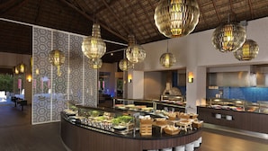 Uma área de buffet com diversas opções de comida, uma cozinha ao fundo e luminárias decorativas penduradas.