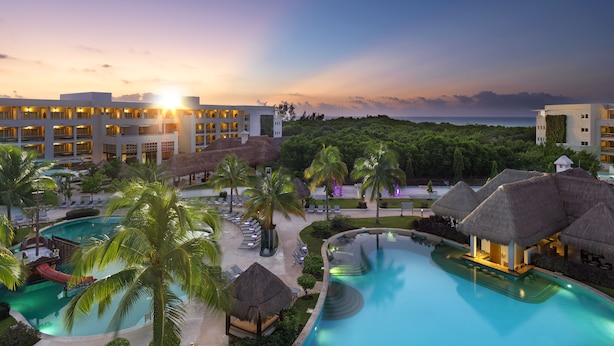 Um resort com várias piscinas, palmeiras e vista para o oceano ao pôr do sol.