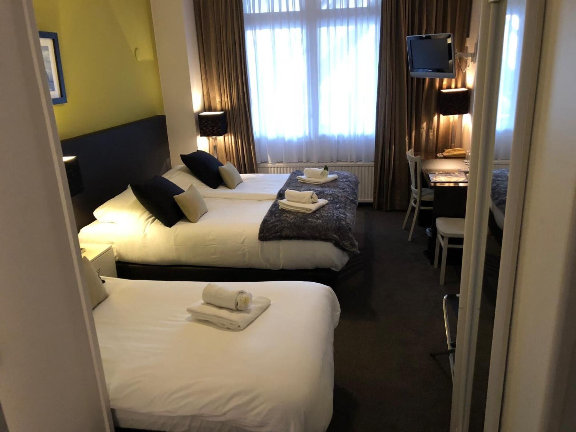 Quarto de hotel com duas camas, uma escrivaninha, uma cadeira, televisão e uma janela com cortinas.