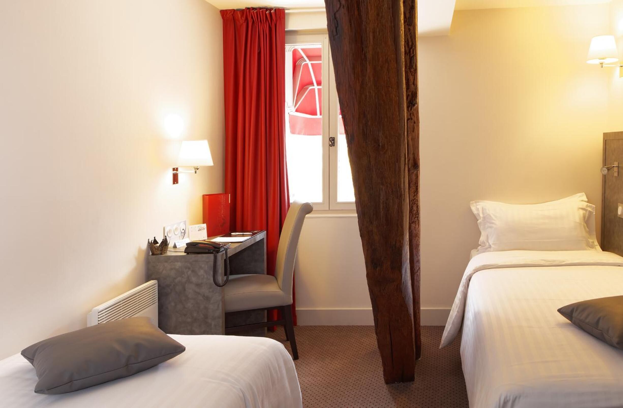Quarto de hotel com duas camas, uma escrivaninha, uma cadeira, um radiador e uma janela com cortinas vermelhas.