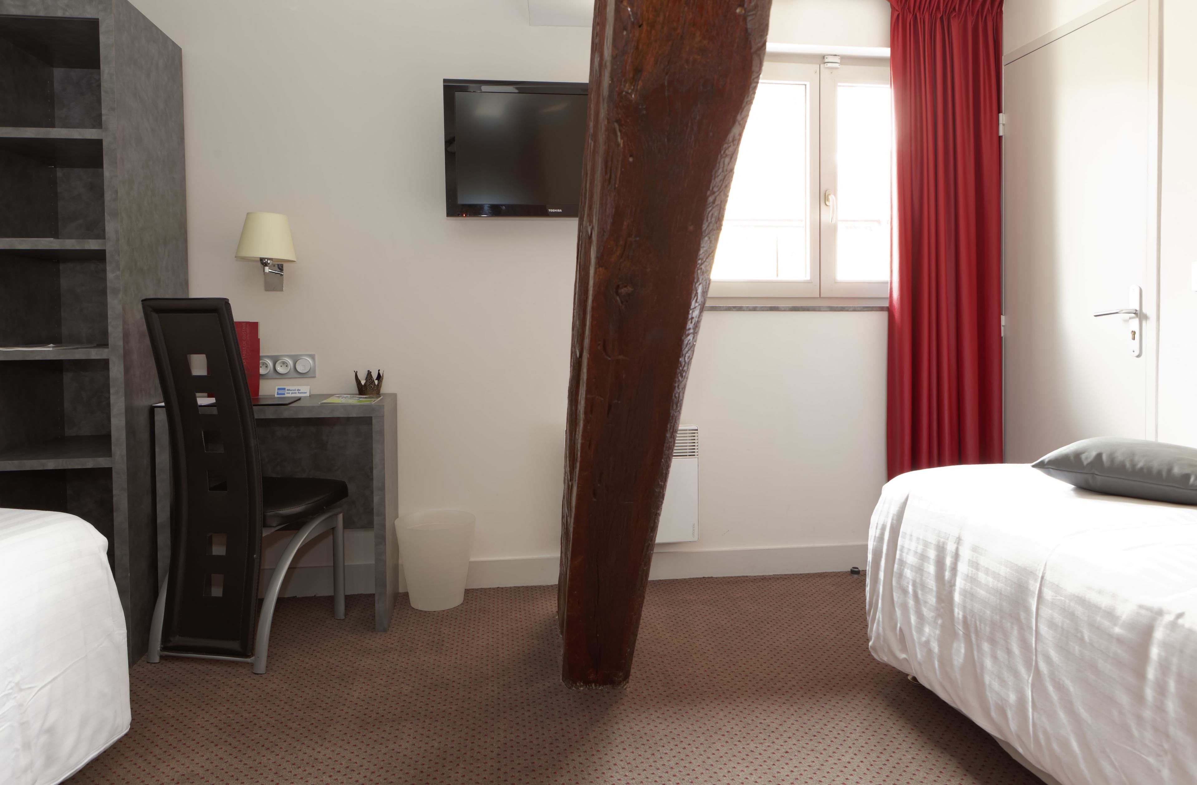 Quarto de hotel com cama, escrivaninha com cadeira, televisão fixada na parede e janela com cortinas vermelhas.