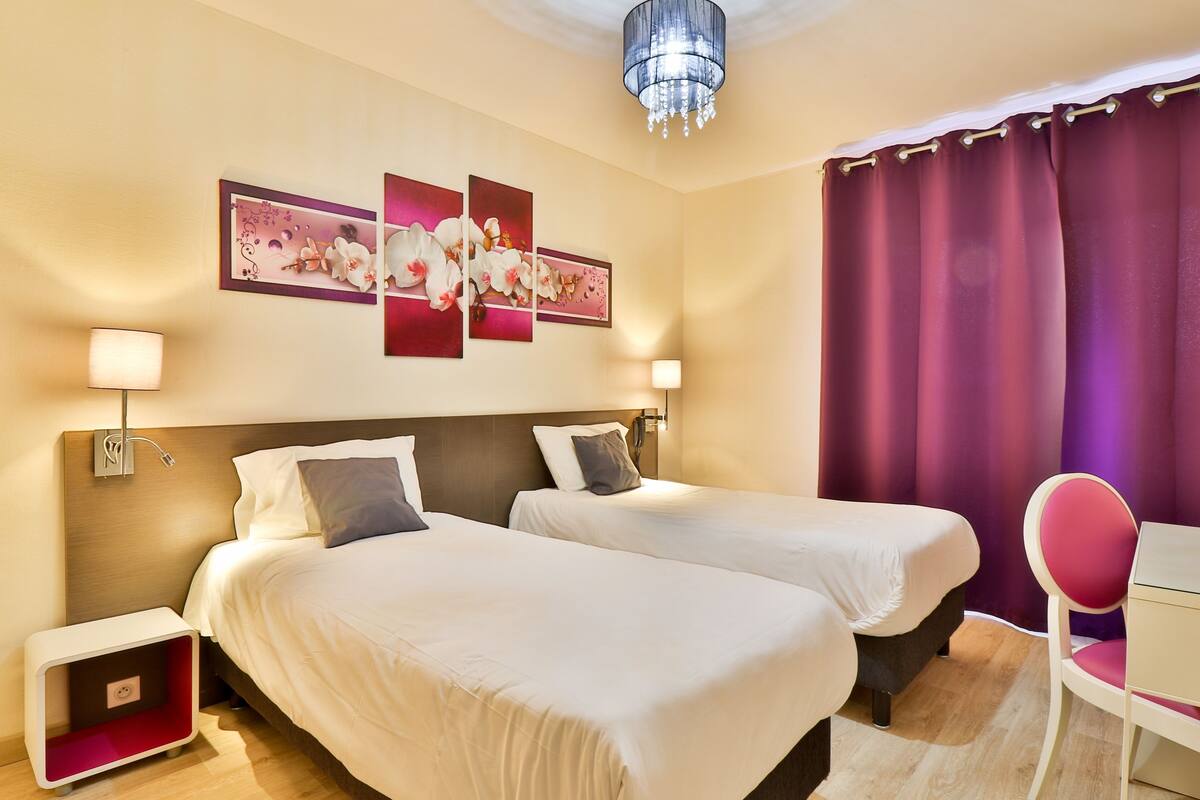 Quarto de hotel com duas camas, uma escrivaninha, uma cadeira e um lustre.
