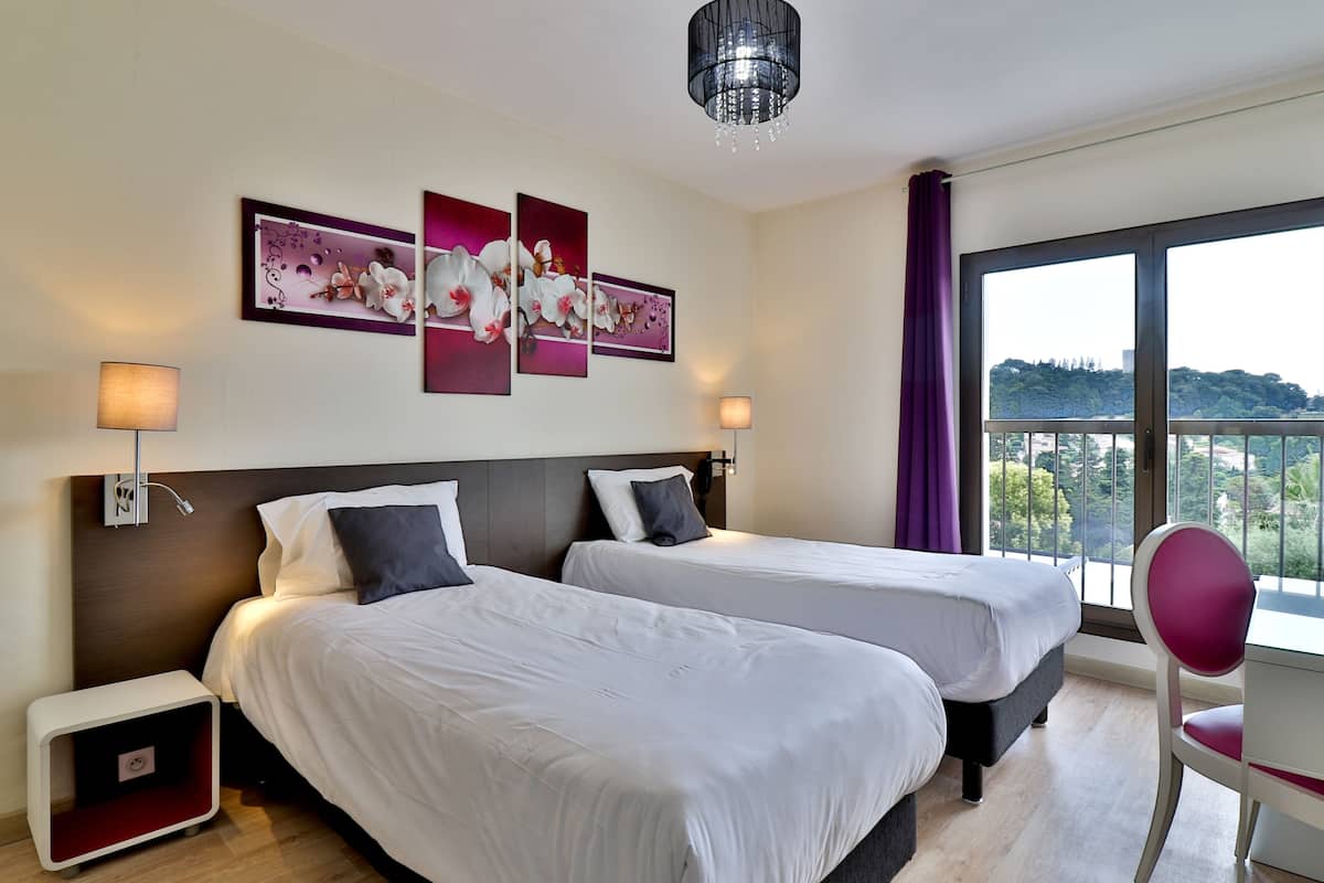 Quarto de hotel com duas camas, uma escrivaninha, uma cadeira e uma janela grande com vista para árvores.