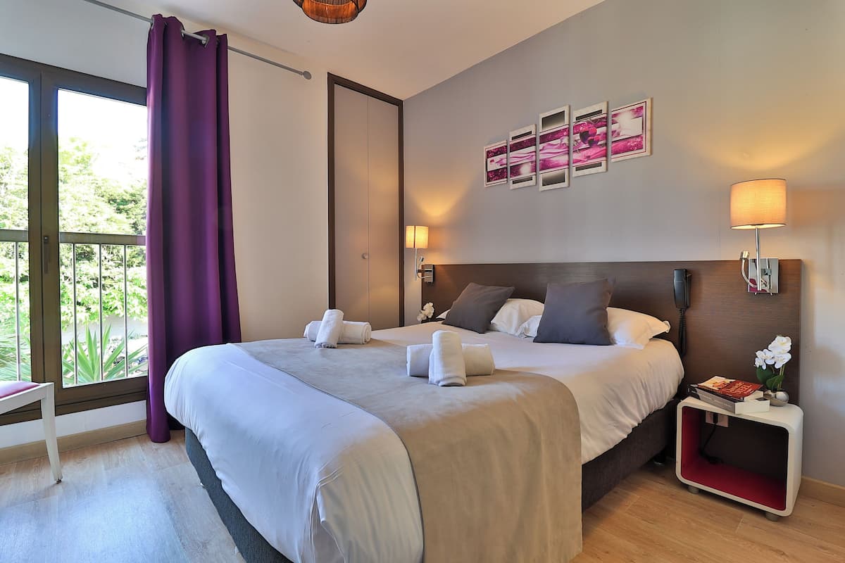 Quarto de hotel moderno com cama grande, duas mesas de cabeceira, um criado-mudo, janela com cortinas roxas e vista para a varanda.