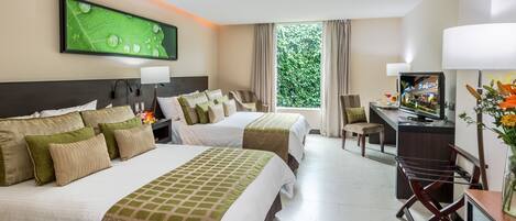 Quarto de hotel com duas camas, uma escrivaninha, uma cadeira, televisão e janela com vista para o verde.
