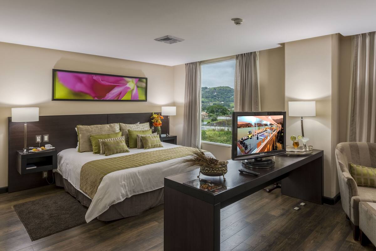 Quarto de hotel com cama, escrivaninha com televisão, cadeira e vista para o exterior.