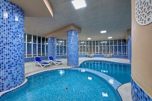 Uma piscina interior com azulejos de mosaico azul, espreguiçadeiras e grandes janelas.