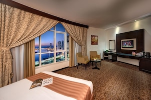 Quarto de hotel com uma janela ampla que oferece vista para a cidade, uma cama com uma revista, uma mesinha com uma tigela, duas poltronas, uma escrivaninha com uma TV e uma cafeteira.