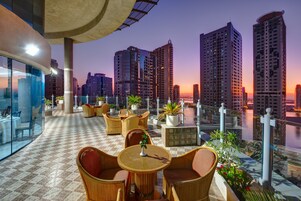 Terraço no terraço com móveis de vime, plantas em vasos e vista para edifícios altos ao pôr do sol.