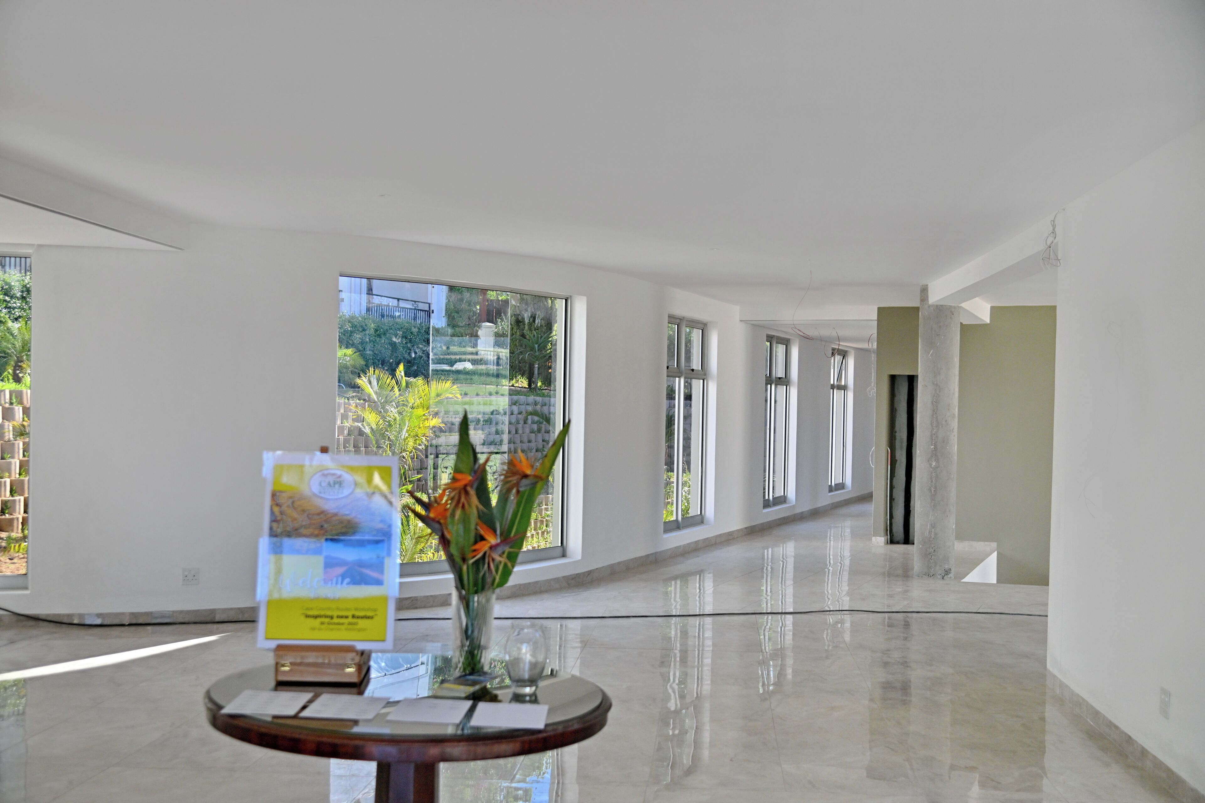 Um hall de hotel espaçoso e moderno, com grandes janelas, piso lustroso e uma pequena mesa com folhetos informativos e um vaso de flores.