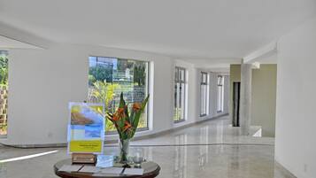 Um hall de hotel espaçoso e moderno, com grandes janelas, piso lustroso e uma pequena mesa com folhetos informativos e um vaso de flores.
