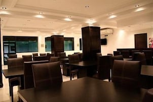 Uma área de refeições moderna com mesas e cadeiras marrons escuras, iluminação embutida no teto e grandes janelas.