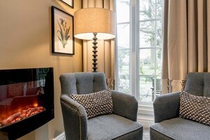 Uma sala de estar aconchegante com dois poltronas cinzas, um abajur, lareira e uma janela grande com cortinas.