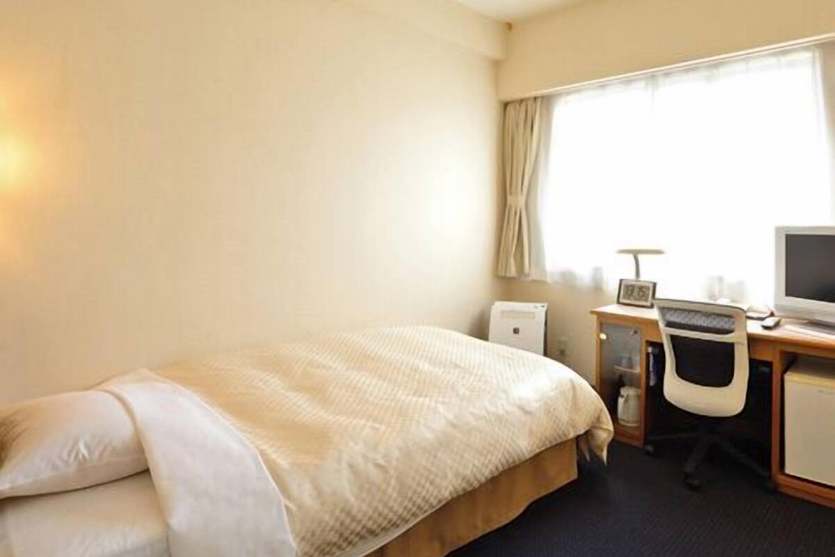 Quarto de hotel com cama, escrivaninha, cadeira, computador e janela.