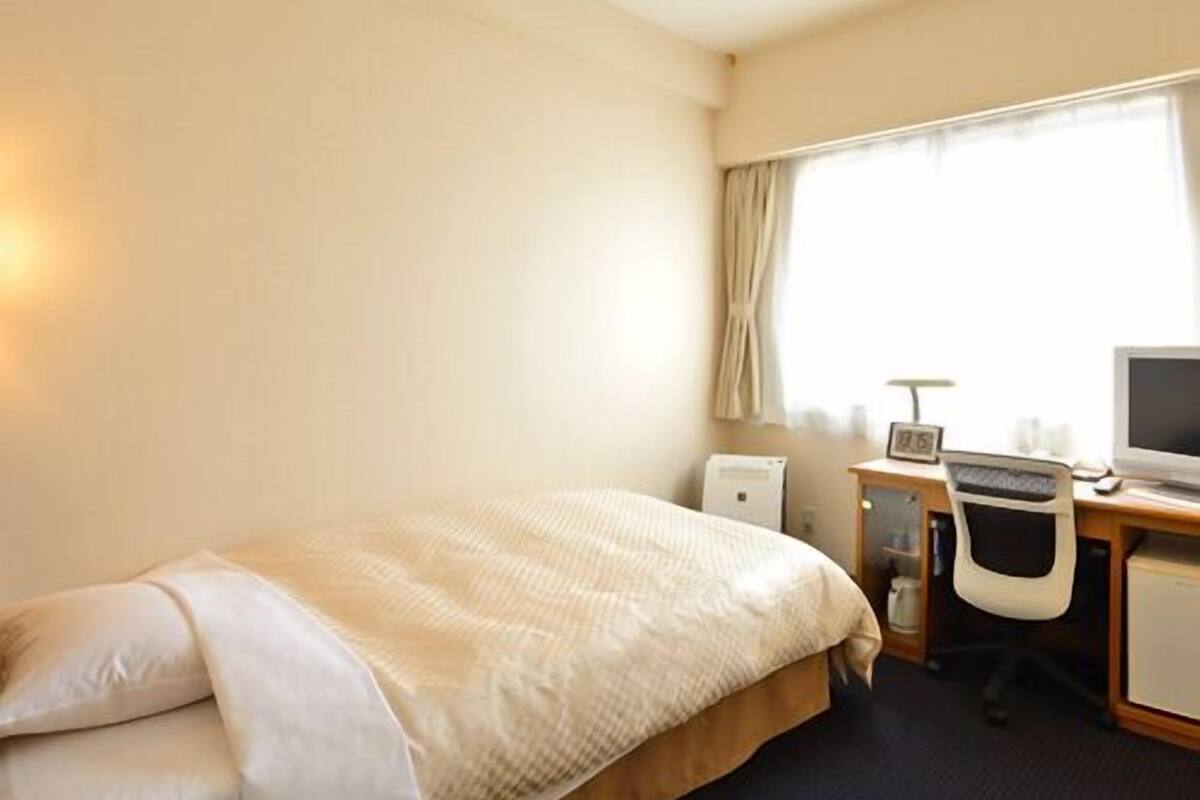 Quarto de hotel com uma cama, uma mesa com computador, uma cadeira e uma janela com cortinas.