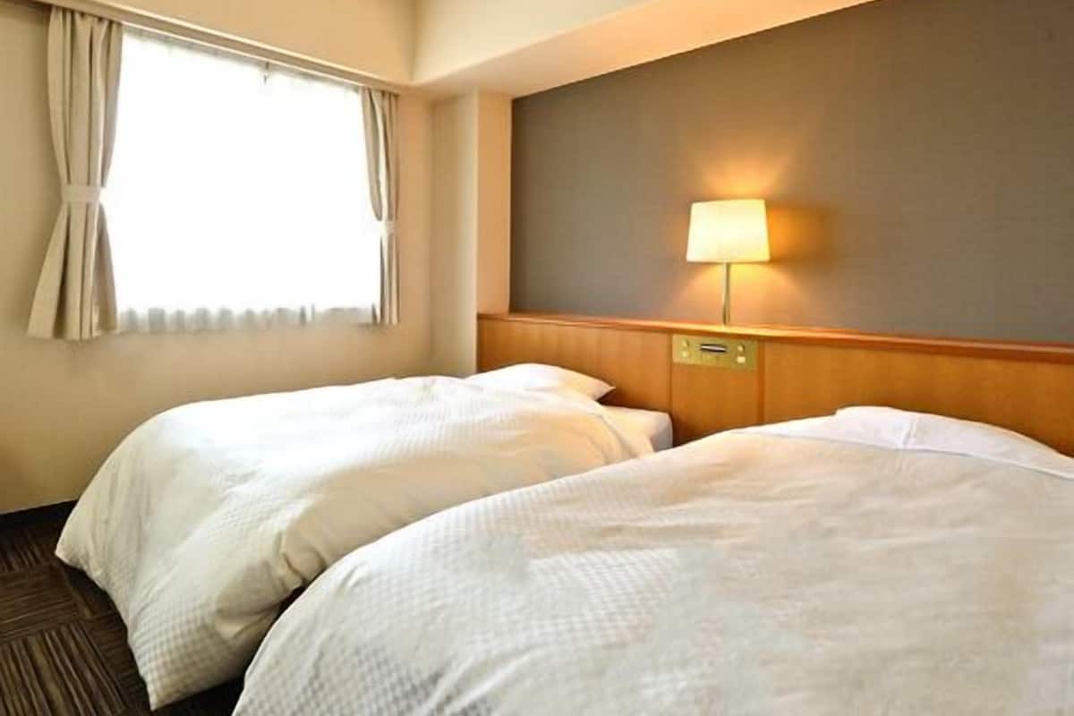 Quarto de hotel com duas camas, uma janela com cortinas, uma luminária na parede e cabeceira de madeira.