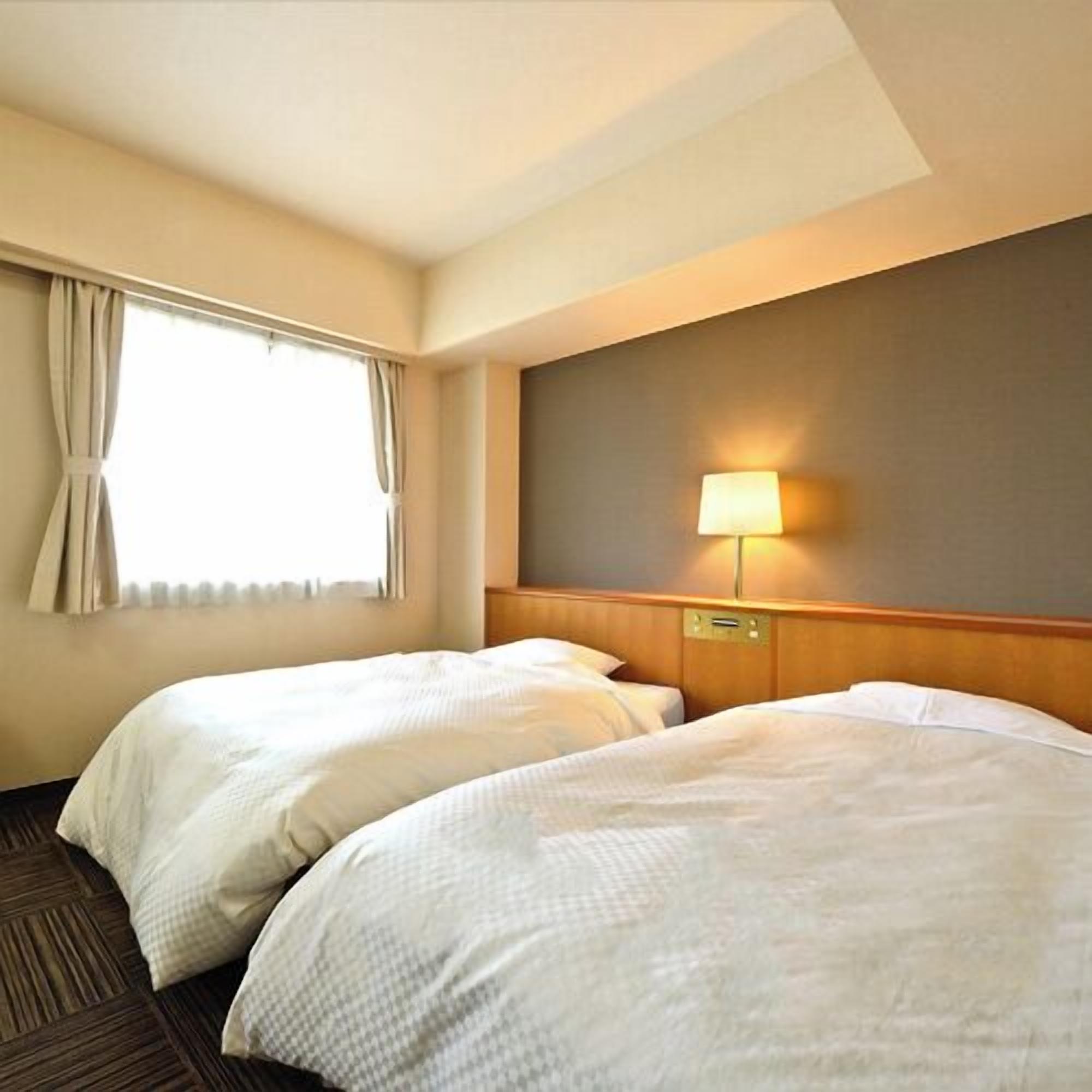 Quarto de hotel com duas camas, uma janela com cortinas, uma luminária fixada na parede e cabeceira de madeira.
