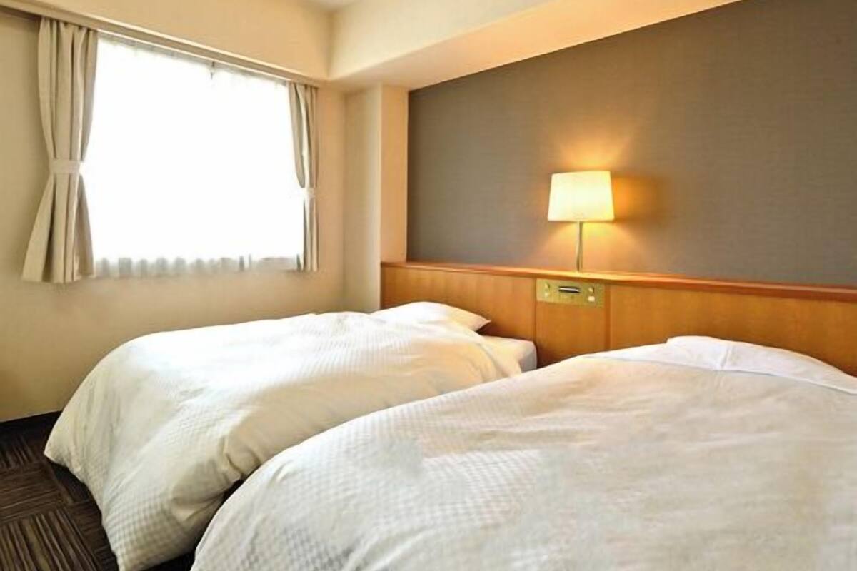 Quarto de hotel com duas camas, uma janela com cortinas, uma luminária fixada na parede e cabeceira de madeira.