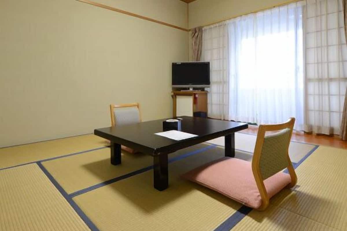 Um espaço com esteiras de tatame, uma mesa baixa, cadeiras, uma televisão e uma janela com cortinas.
