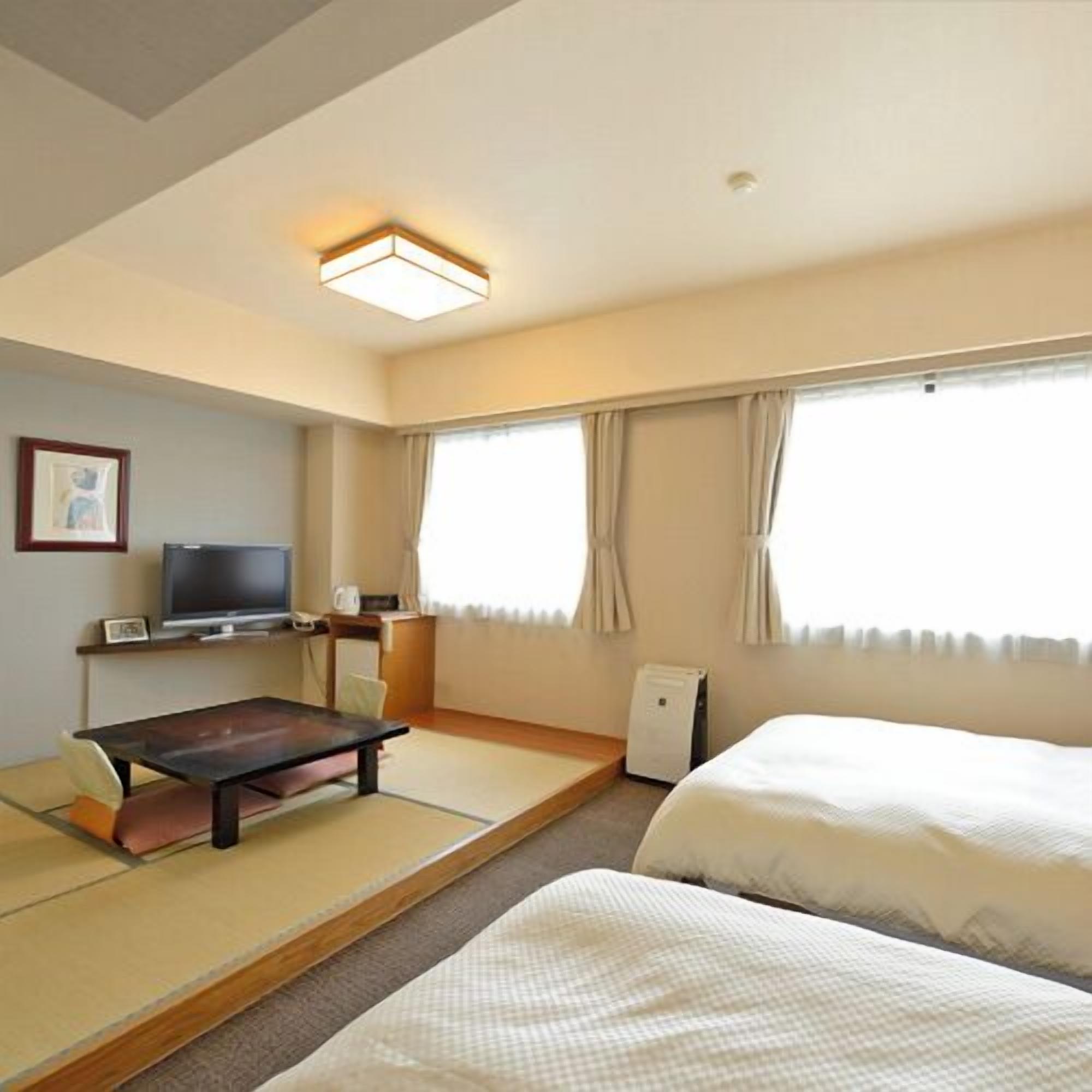 Quarto de hotel com cama, escrivaninha, televisão e janela com cortinas.