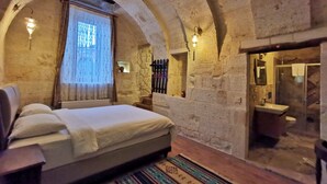 Quarto com teto abobadado de paredes de pedra, cama com lençóis brancos, soalho de madeira e banheiro com pia e espelho.