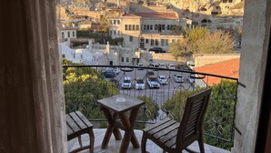 Varanda com duas cadeiras de madeira e uma pequena mesa, com vista para um estacionamento e edifícios ao fundo de montanhas.