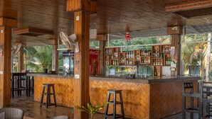 Uma área de bar com balcão de madeira, banquetas, um anúncio de Coca-Cola e prateleiras com garrafas e copos.