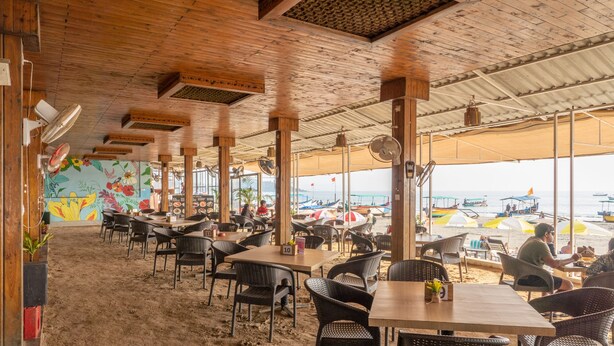 Restaurante ao ar livre com mesas e cadeiras de madeira, um mural na parede e vista para a praia e barcos.
