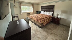 Quarto de hotel com cama, escrivaninha, cadeira, janela com cortinas, televisão fixada na parede e mesa de cabeceira com abajur.