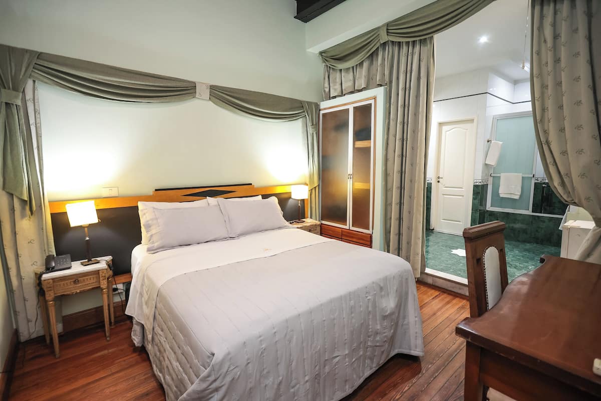 Quarto de hotel com cama, mesa de cabeceira, abajur, cadeira de madeira e guarda-roupa.