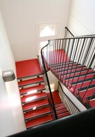Uma escada com degraus cobertos por carpete vermelho e corrimãos pretos, conduzindo a um patamar com uma porta branca.